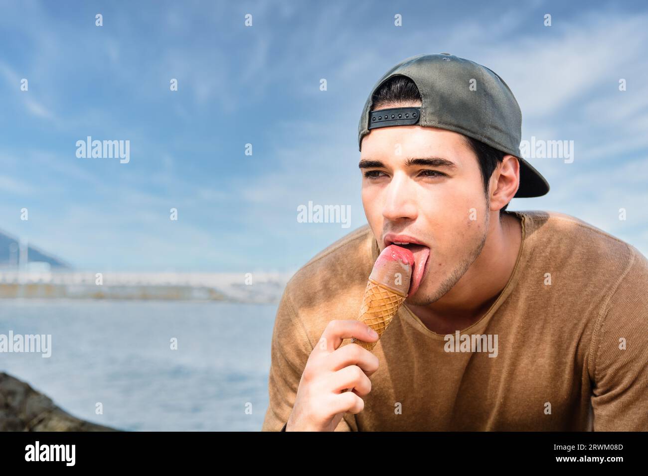 A man with a hat on eating an ice cream cone at the seaside, by the sea ...
