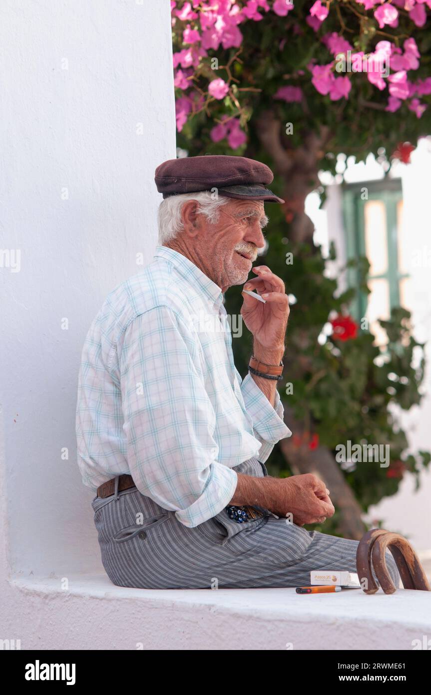 Older Greek man smokes a cigarette while sitting on a white wall and ...
