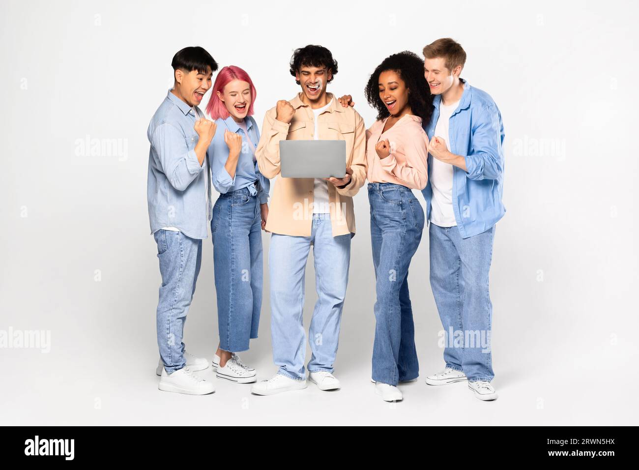 Group of overjoyed multiracial students celebrating success, holding laptop and clenching fists ...