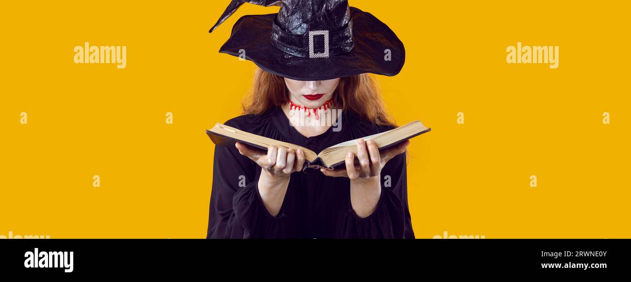 Young woman dressed as evil witch hiding her face under hat and reading ...