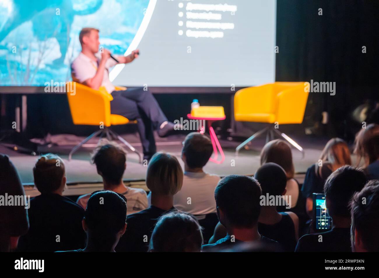 Crowd sitting in hall against blurred stage, while speaker showing ...