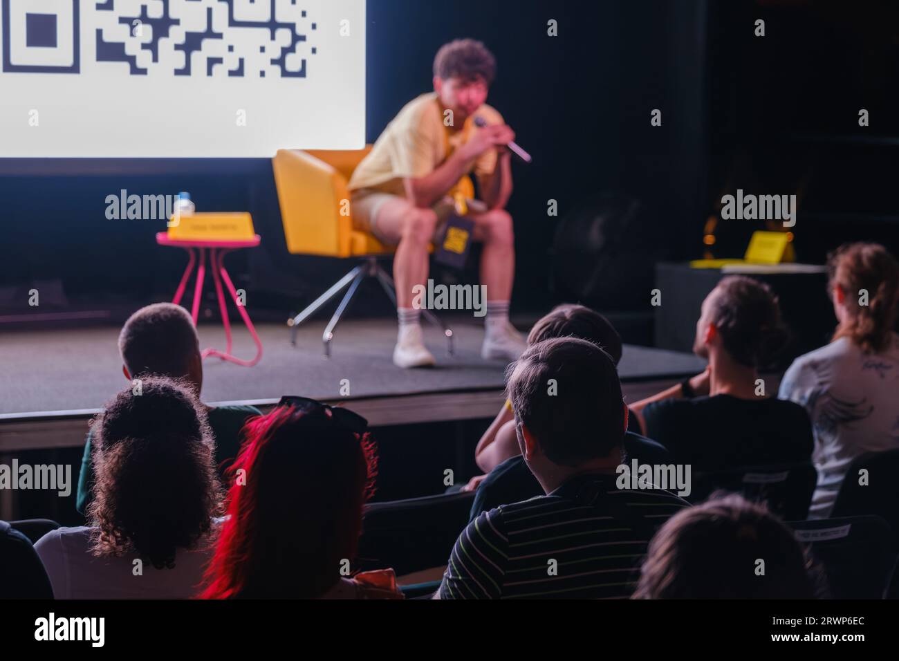 Blurred male coach sitting on chair on stage, near screen and answering ...