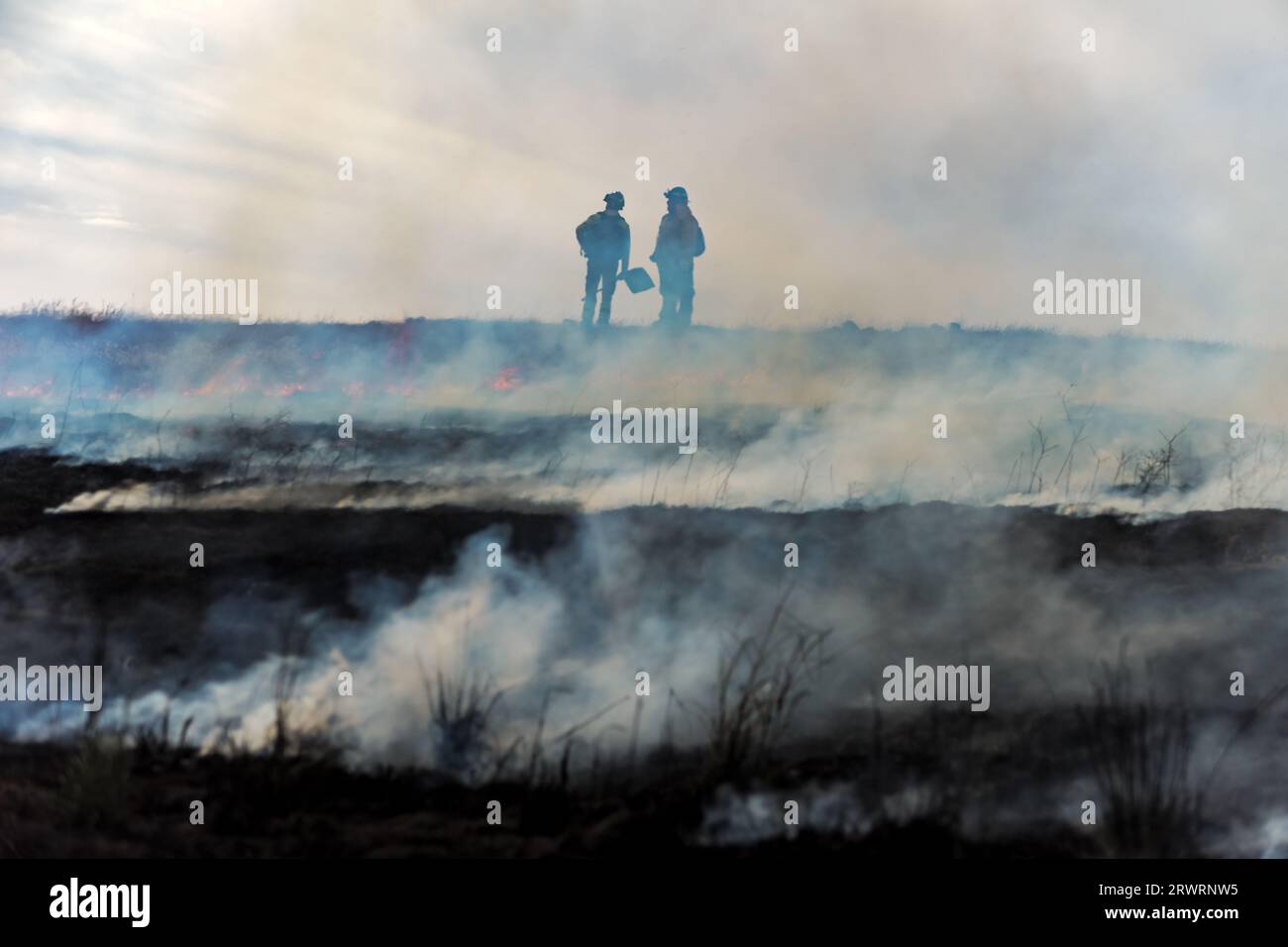 Two firefighters stand silhouetted in smoke on a smoldering hill after ...