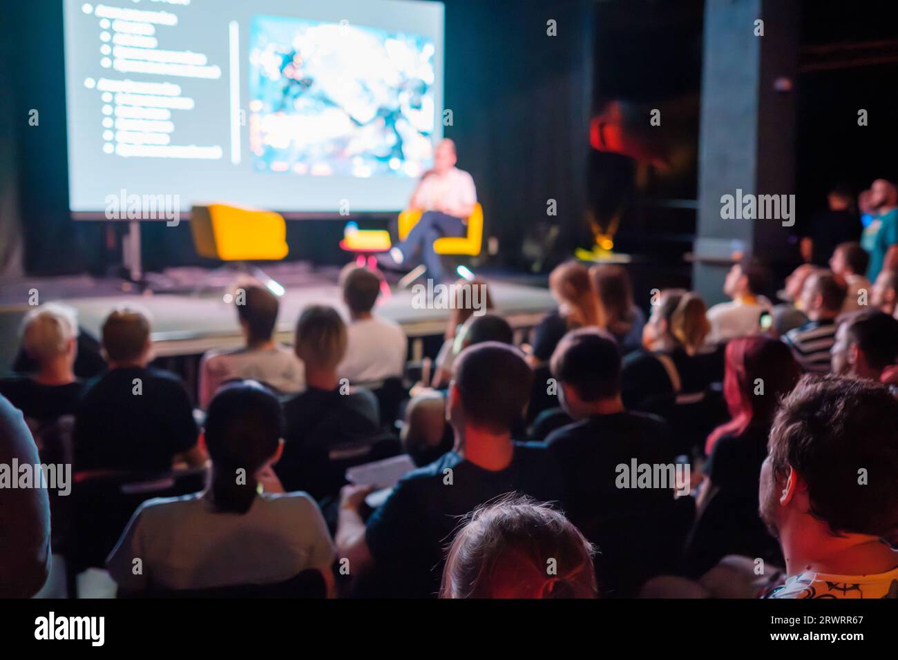 Crowd sitting in hall against blurred stage, while speaker showing ...