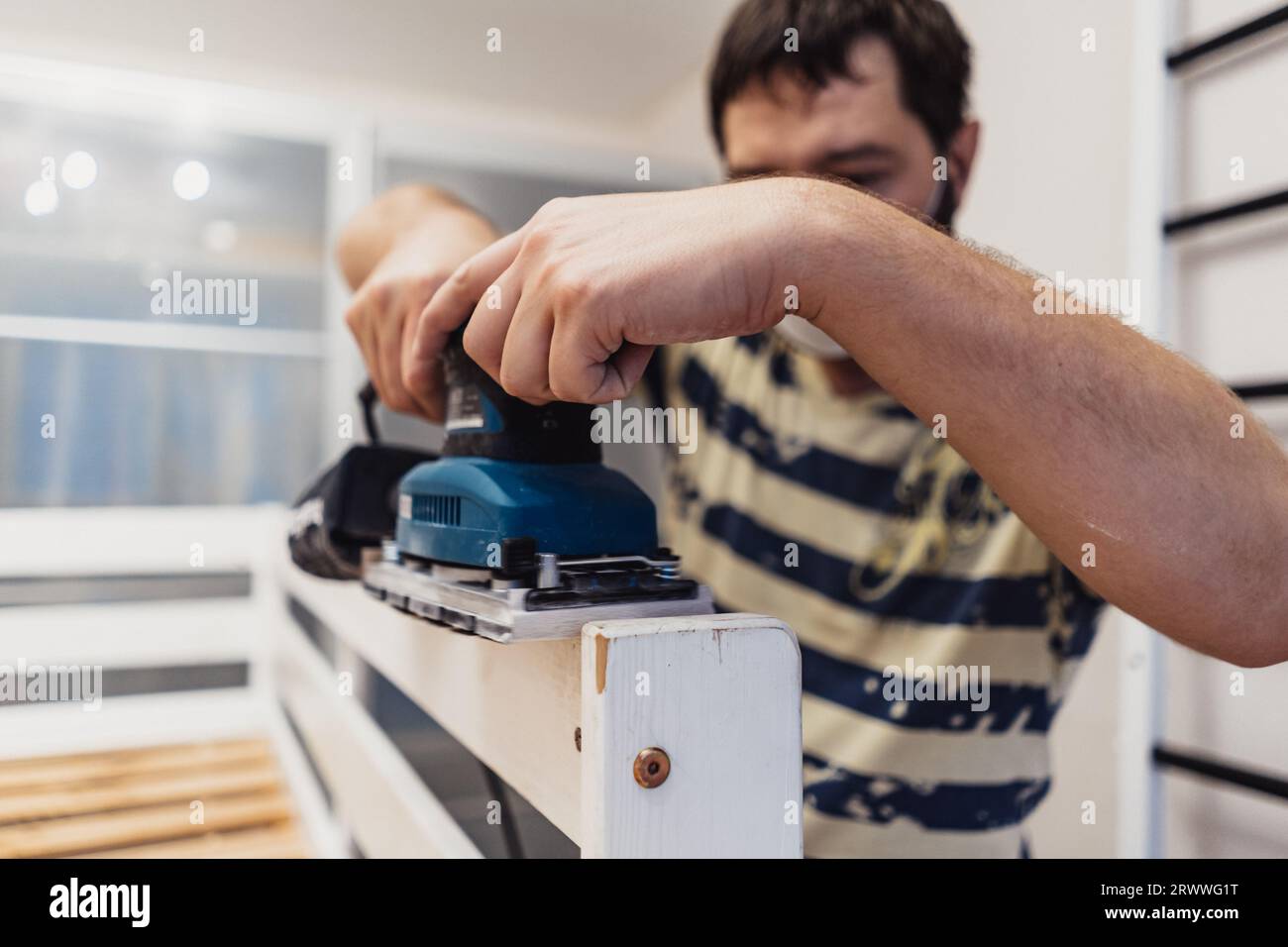 man wearing protective mask rubs wooden child bed with power sander or ...