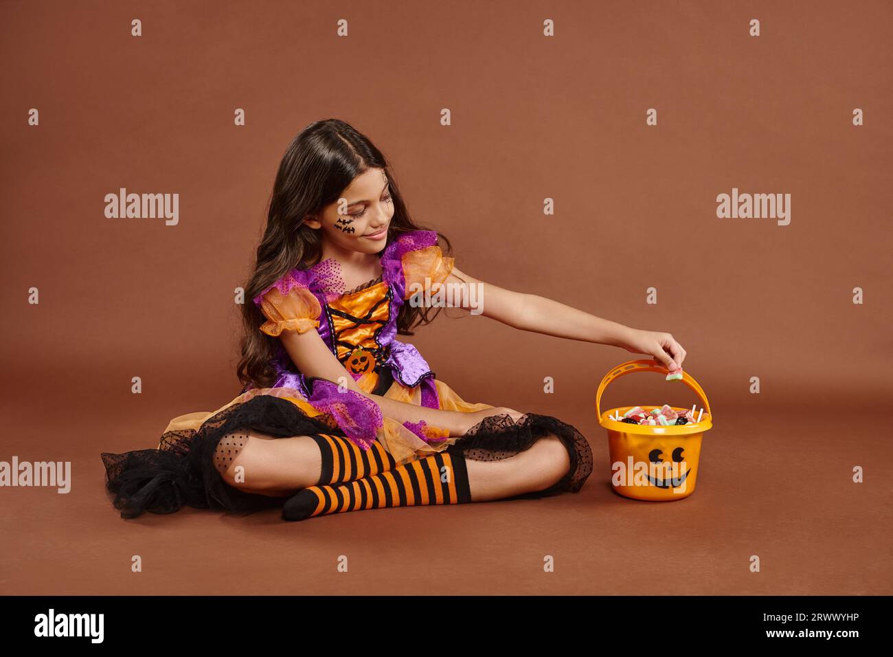 positive girl in colorful Halloween costume sitting near bucket with ...