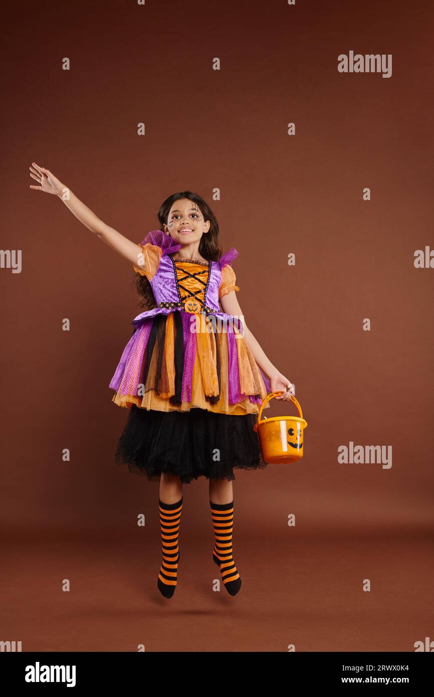 happy girl in Halloween costume levitating with candy bucket on brown ...