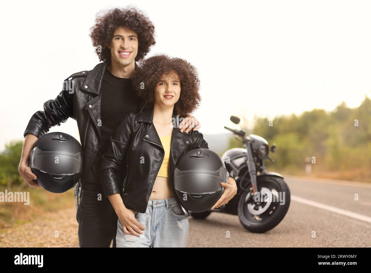 Couple in leather jackets holding helmets in front of a parked motorbike on an open road Stock ...