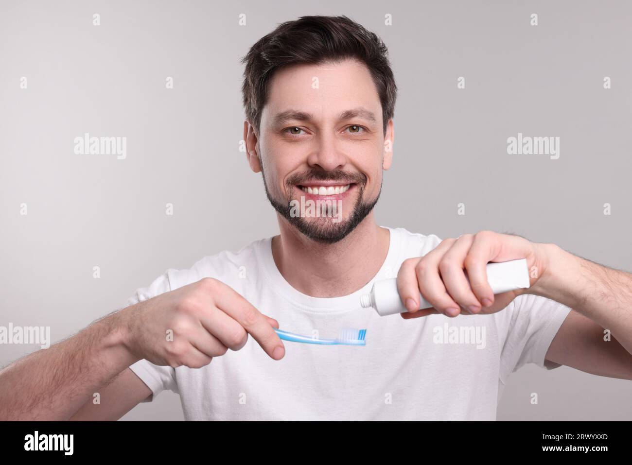 Happy man squeezing toothpaste from tube onto toothbrush on light grey ...
