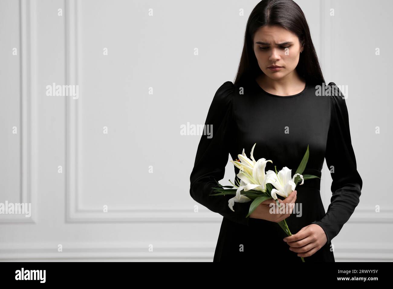 Sad woman with lilies mourning near white wall, space for text. Funeral ...