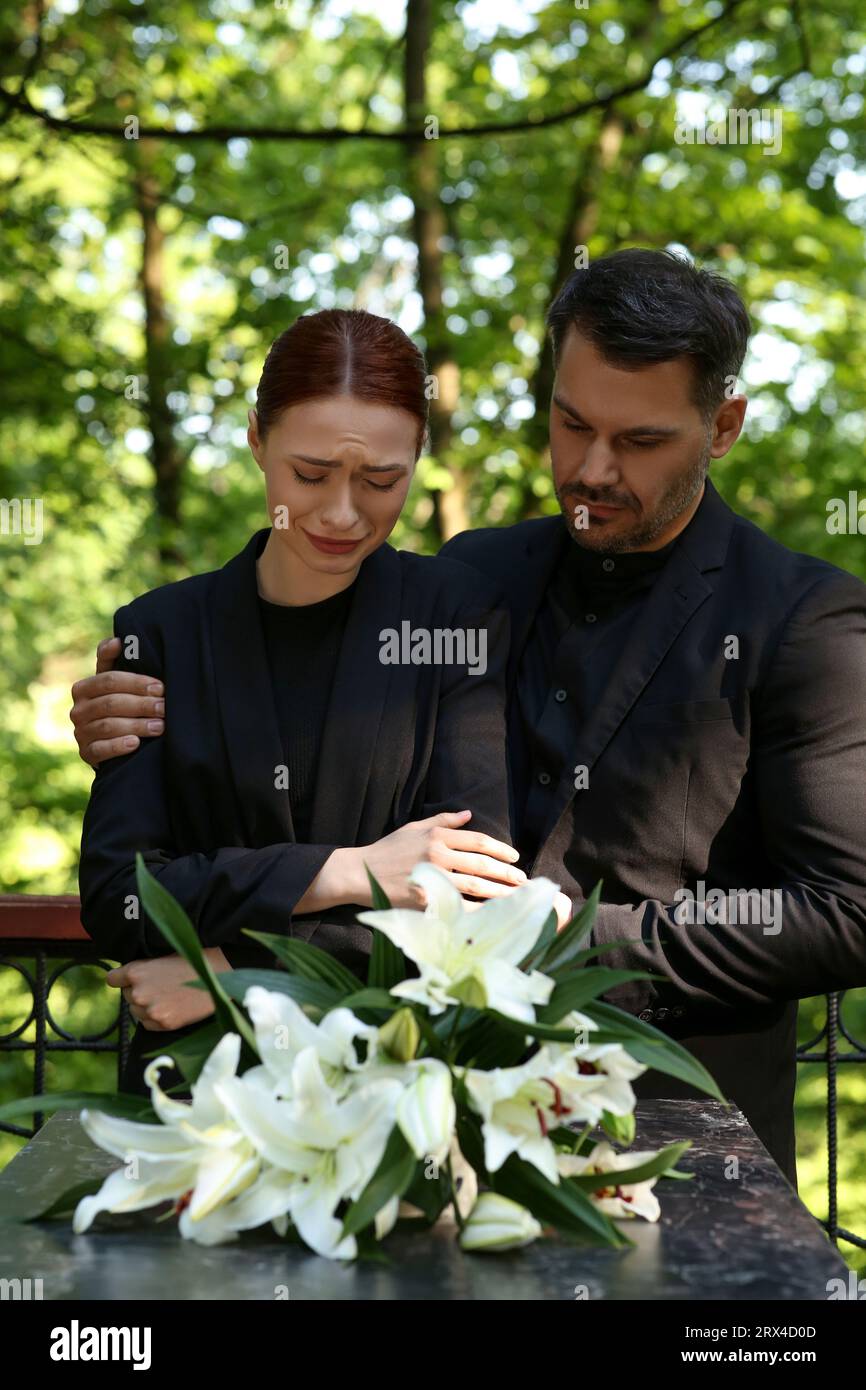 Sad couple mourning near granite tombstone with white lilies at ...