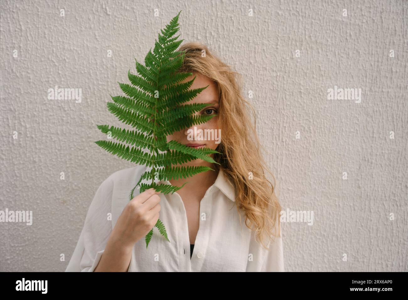 Young woman covering half face with fern leaf in front of wall Stock ...