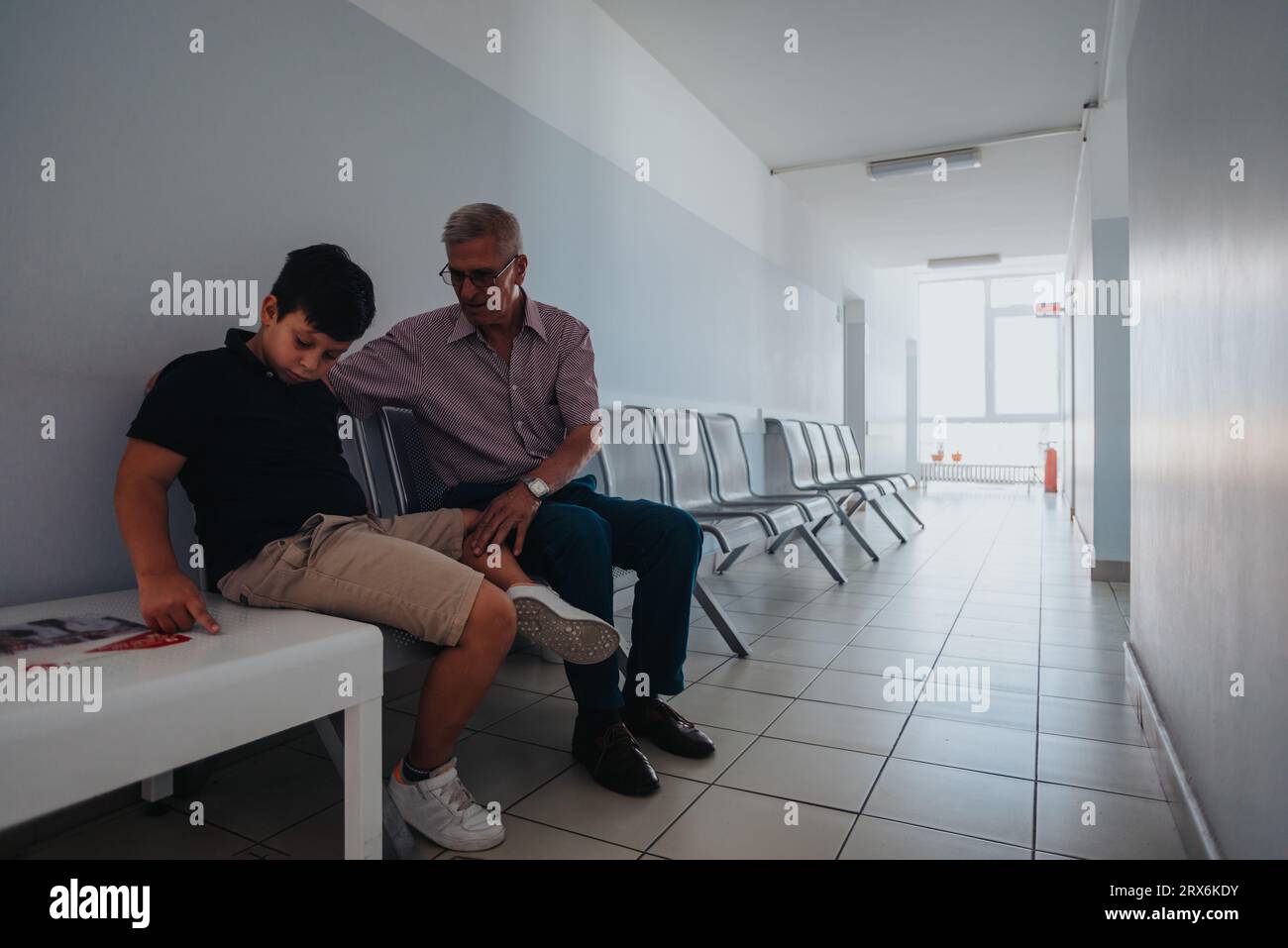 Child with his grandfather awaiting their turn in a hospital while sitting on a bench in the ...