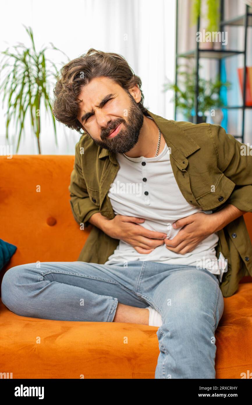 Middle eastern man sits on sofa feeling sudden strong abdominal stomach ...