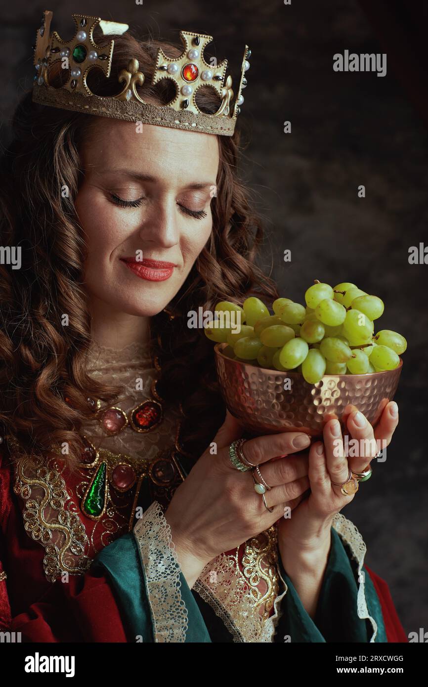 medieval queen in red dress with plate of grapes and crown on dark gray ...