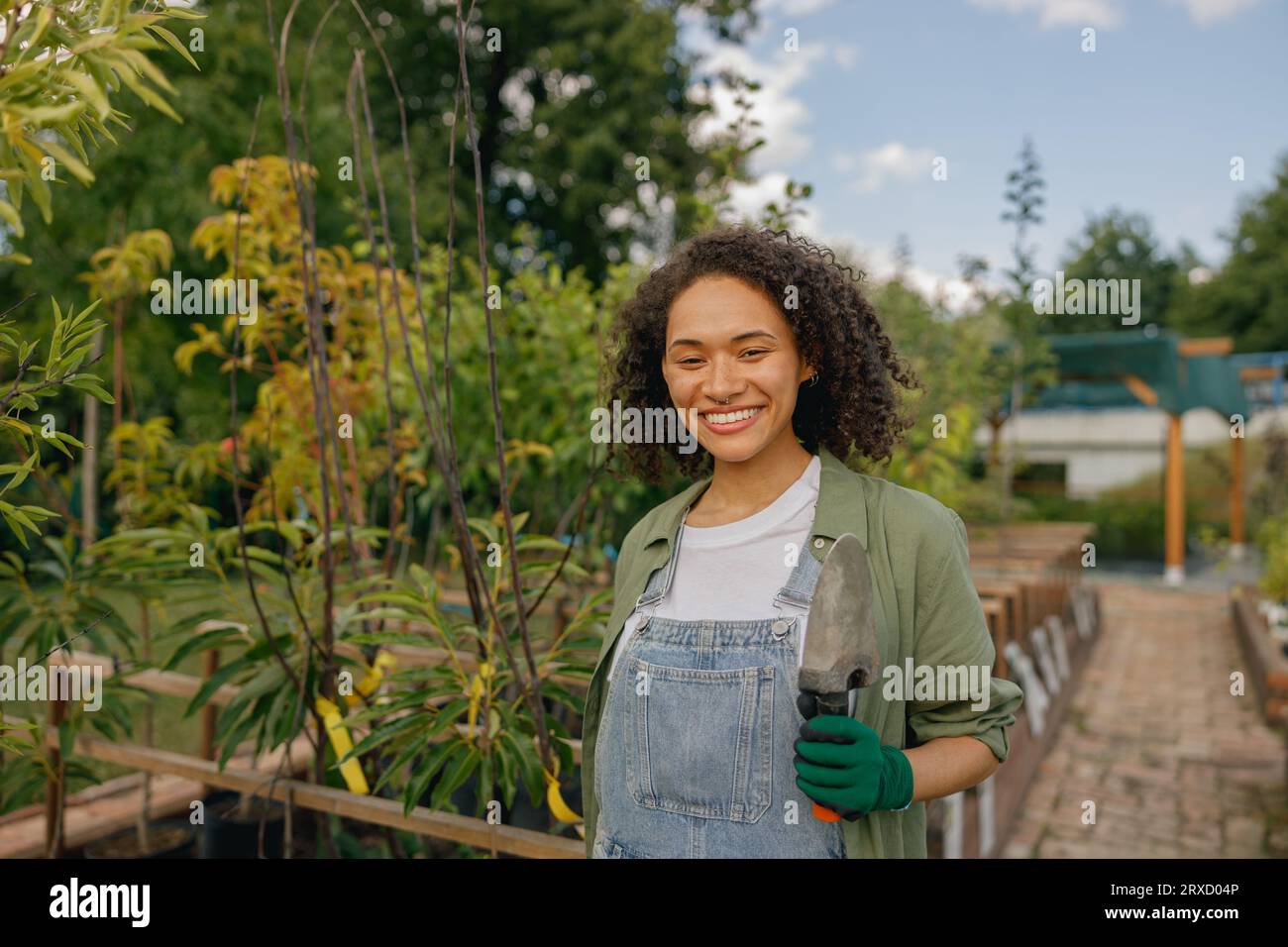 Female gardener standing with hand trowel and looks camera on plants ...