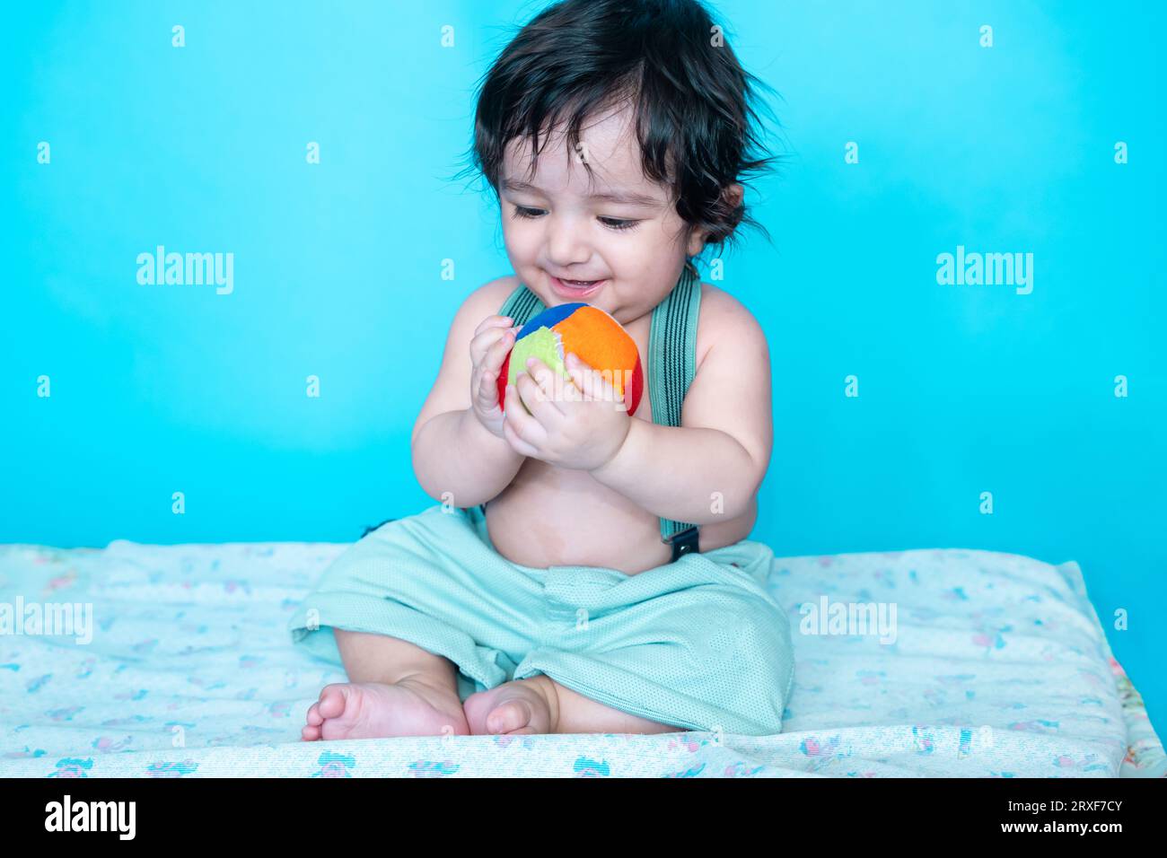 Smiling six months old cute indian baby boy play with toy sitting isolated over blue studio ...