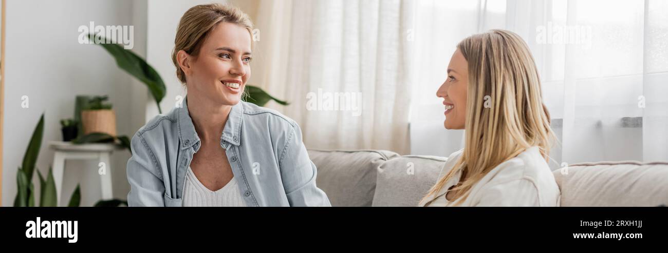 two pretty charming sisters in casual attire sitting on sofa looking at ...