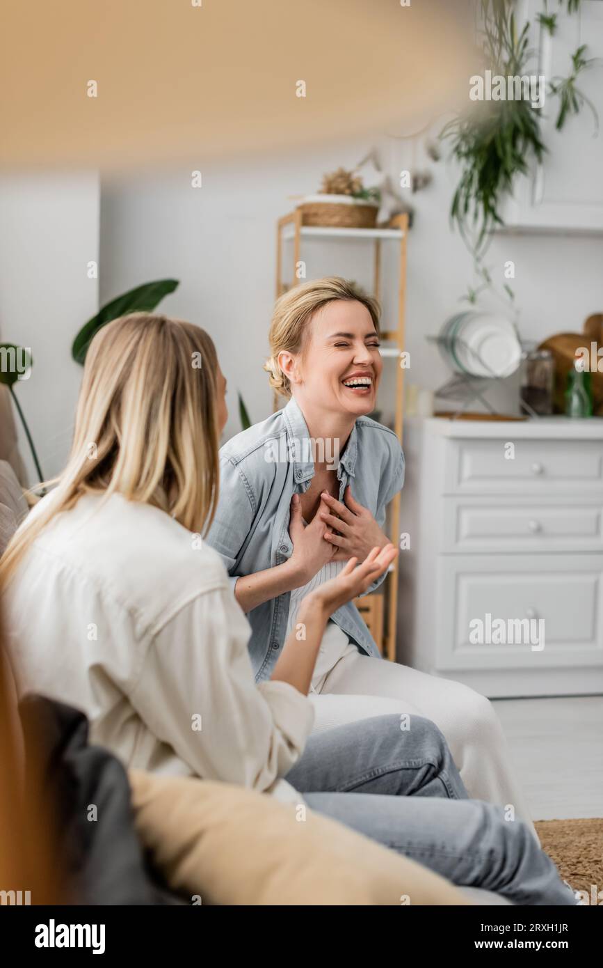 two attractive sisters in casual clothing sitting on sofa, talking and laughing, family bonding ...