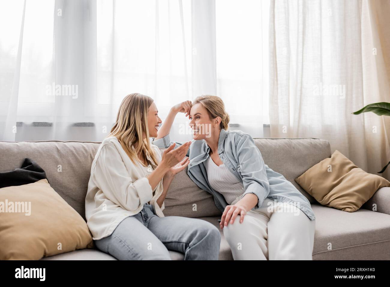 two cheerful lovely sisters sitting on sofa smiling and talking to each other, family bonding ...