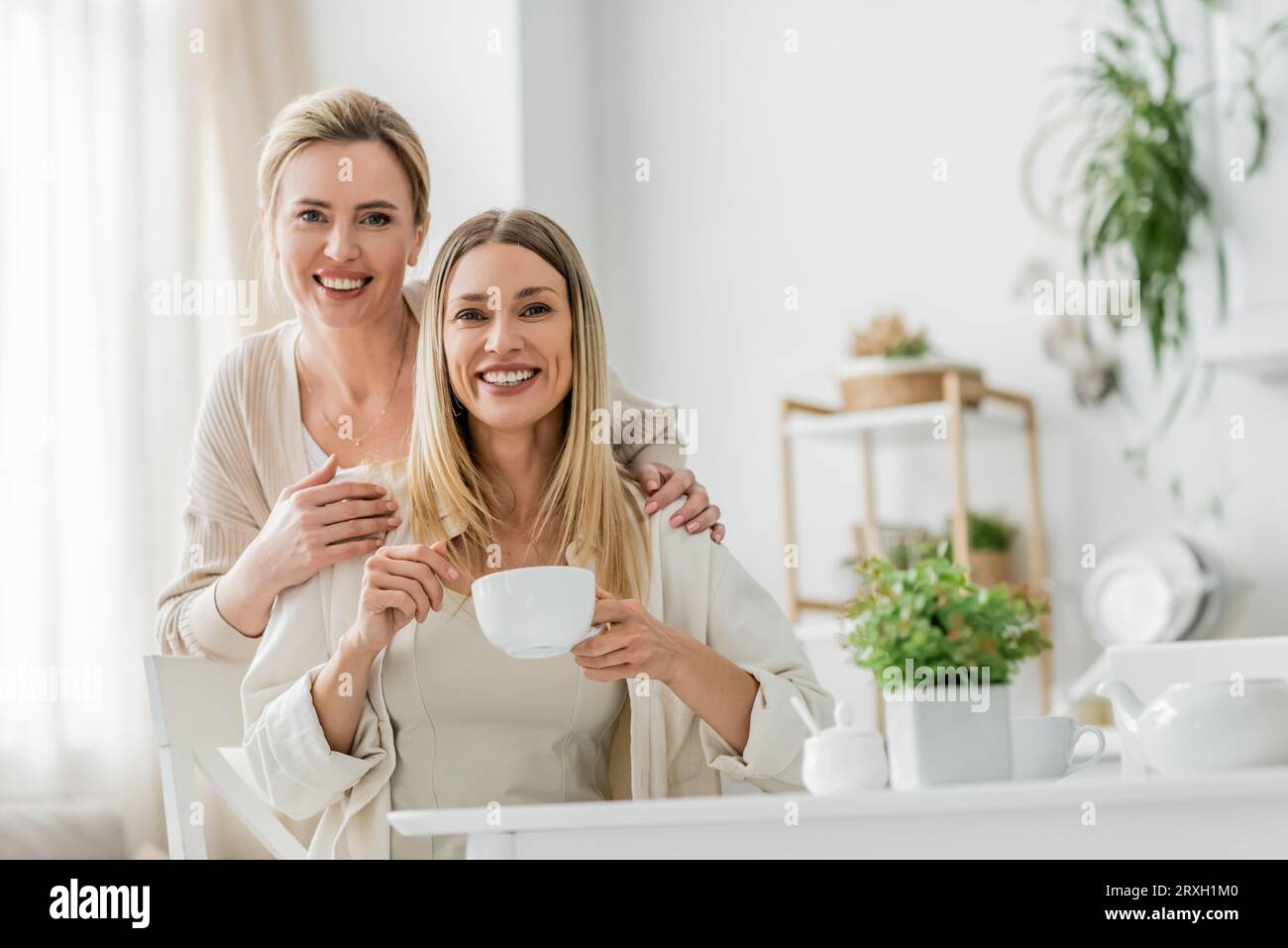 two cheerful pretty sisters looking at camera smiling and posing on kitchen backdrop, family ...