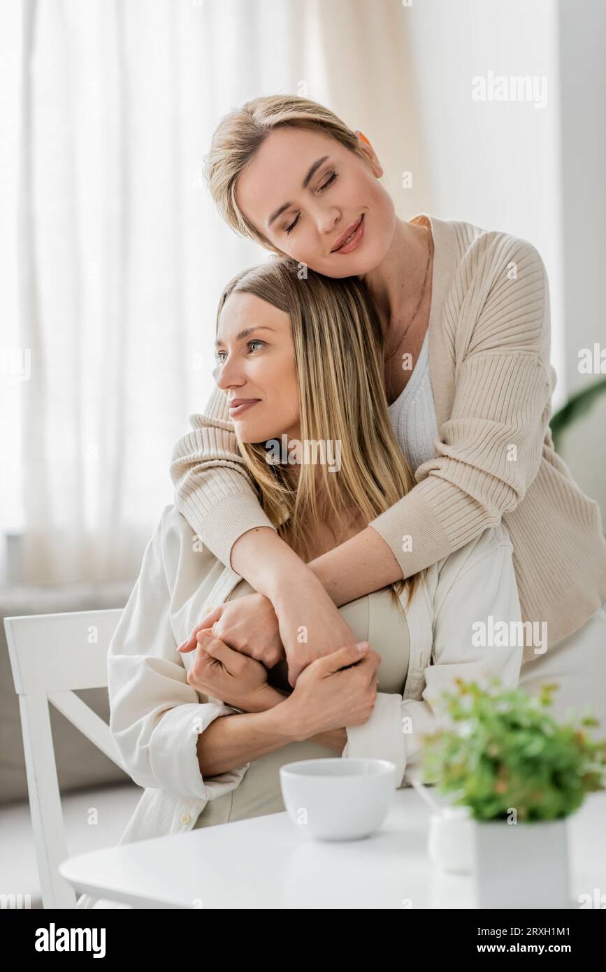 portrait of attractive blonde sisters hugging and holding hands on kitchen backdrop, family ...