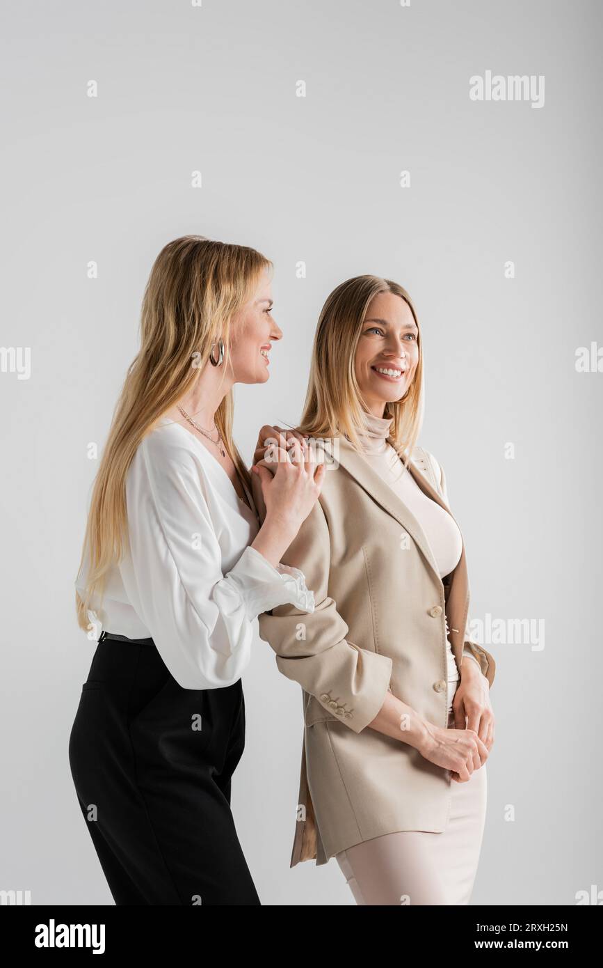 two cheerful attractive blonde sisters smiling and posing on grey backdrop, fashion concept ...
