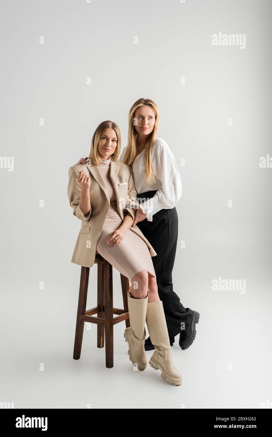 two elegant stylish sisters posing on grey backdrop one sister sitting on chair, bonding ...