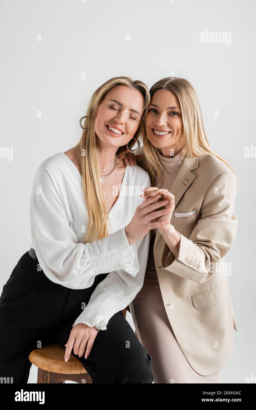 two attractive elegant sisters sitting on chairs smiling and holding hands, fashion and style ...