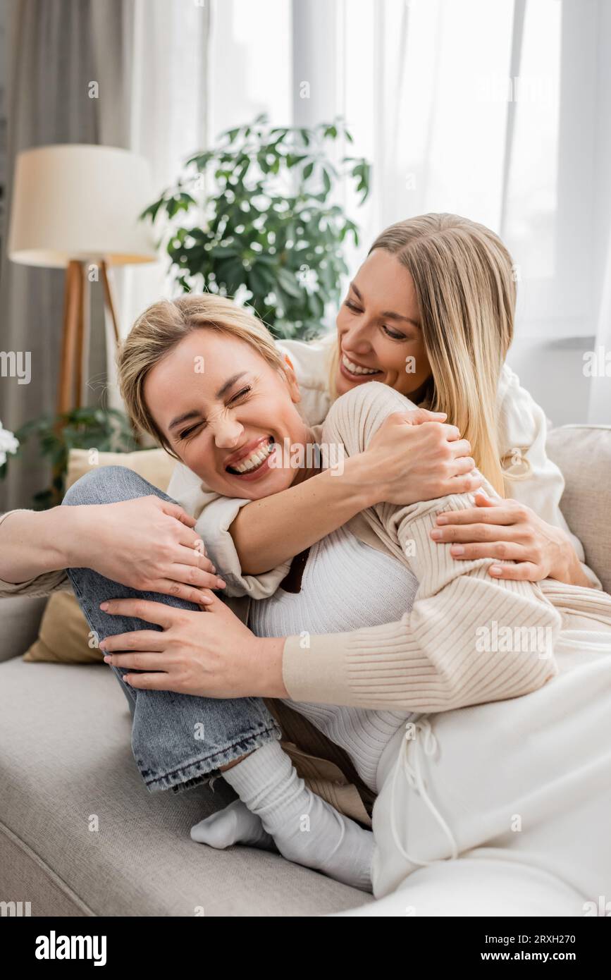 two attractive lovely sisters on sofa laughing and hugging warmly, togetherness, family bonding ...