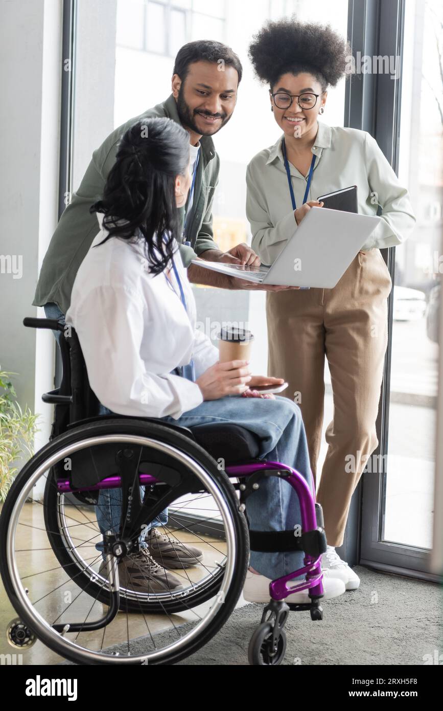 happy interracial colleagues looking at disabled coworker on wheelchair ...