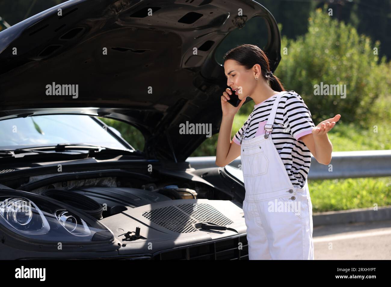 Stressed woman talking on smartphone while looking under hood of broken car outdoors Stock Photo ...