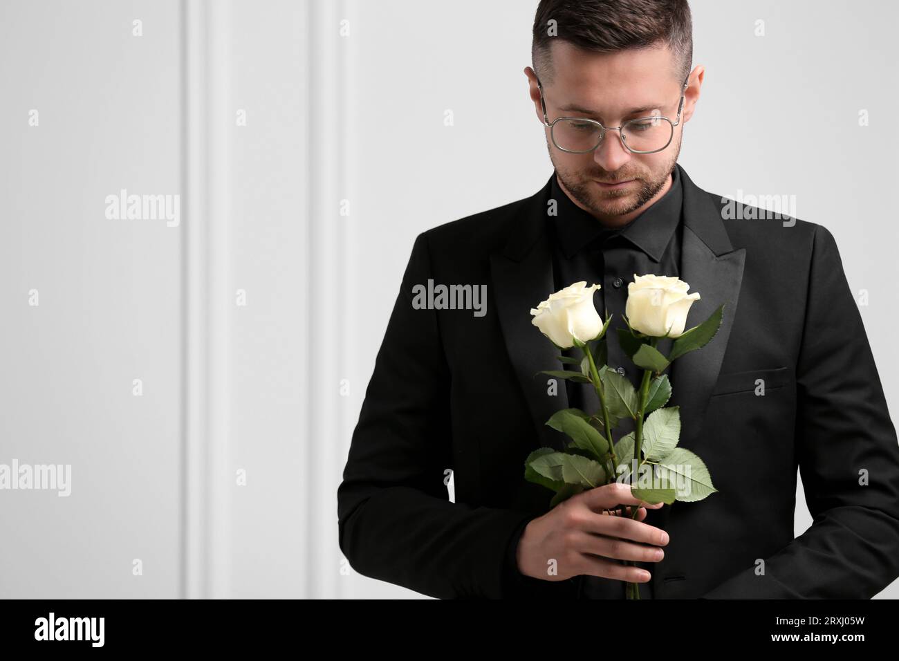 Sad man with rose flowers near white wall, space for text. Funeral ...