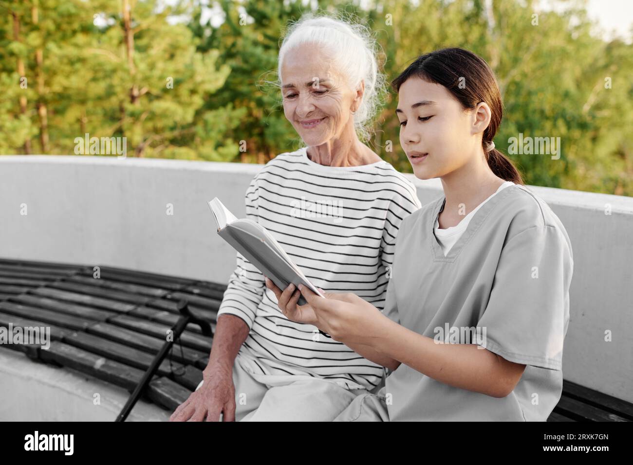 Nurse reading book to senior woman while they sitting on bench outdoors ...