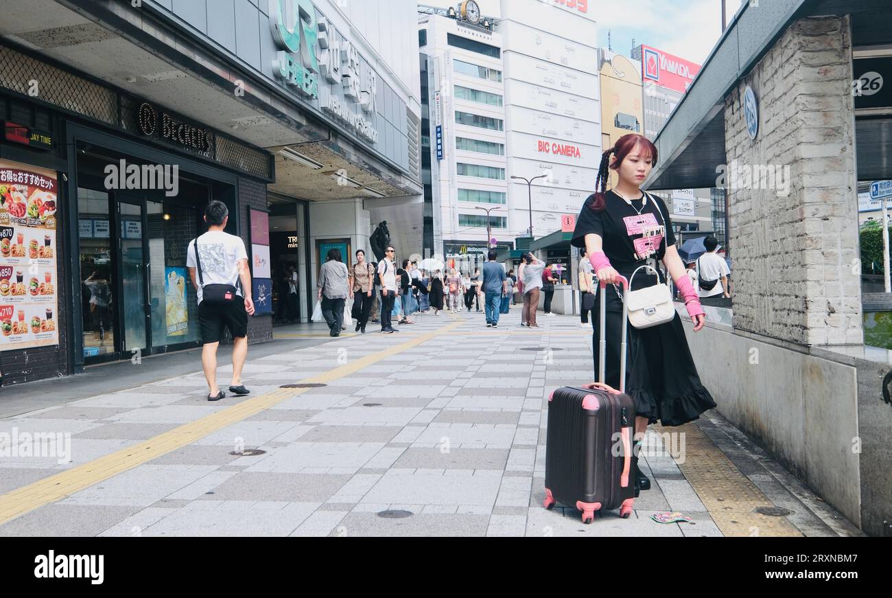 A Japanese girl with a small suitcase stands on the pavement in ...
