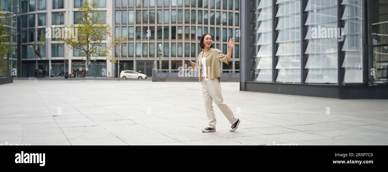 Happy asian girl saying bye to someone, waving at friend and walking on ...