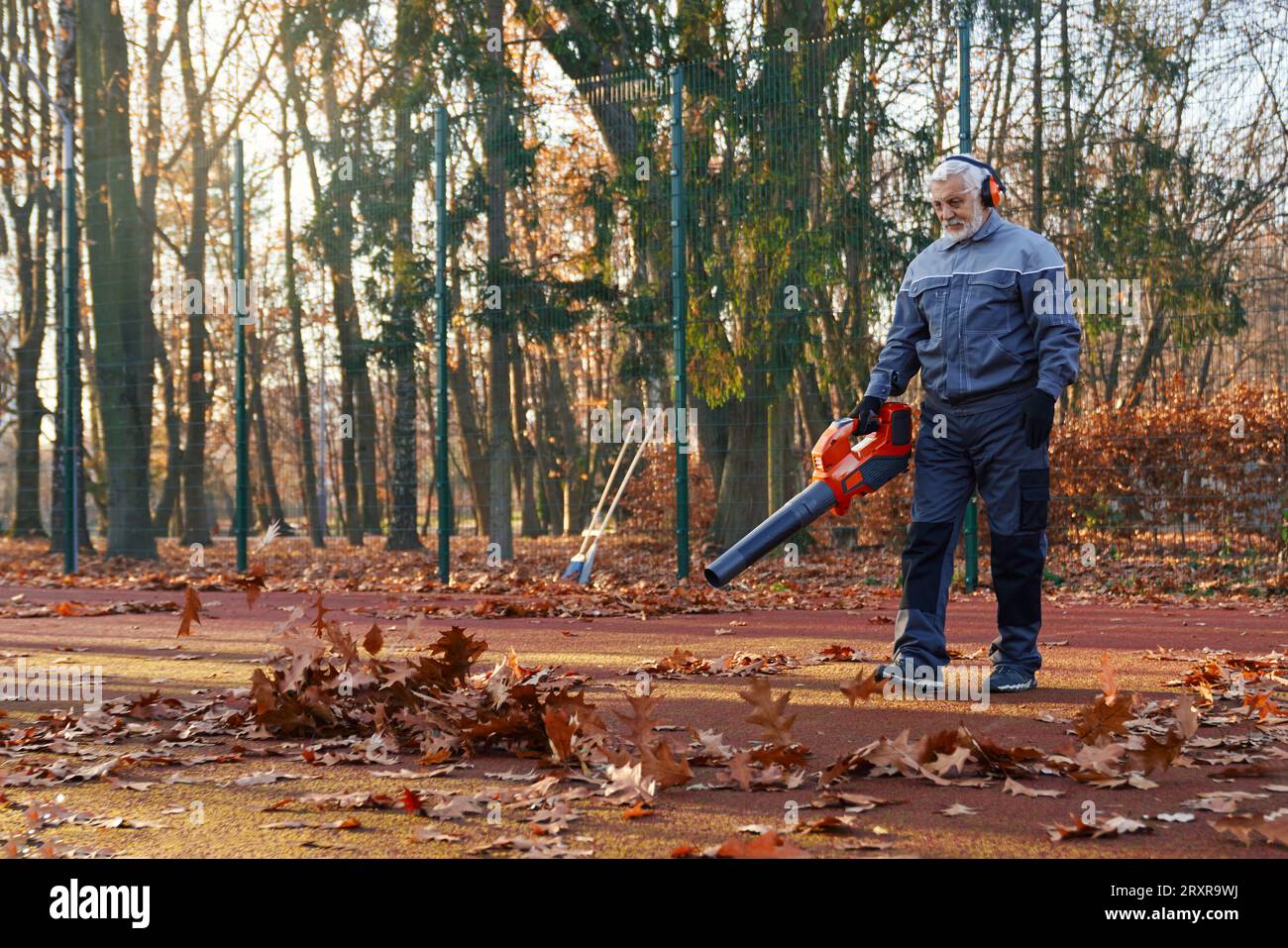 Senior man in overalls, headphones and gloves cleaning fenced city park ...