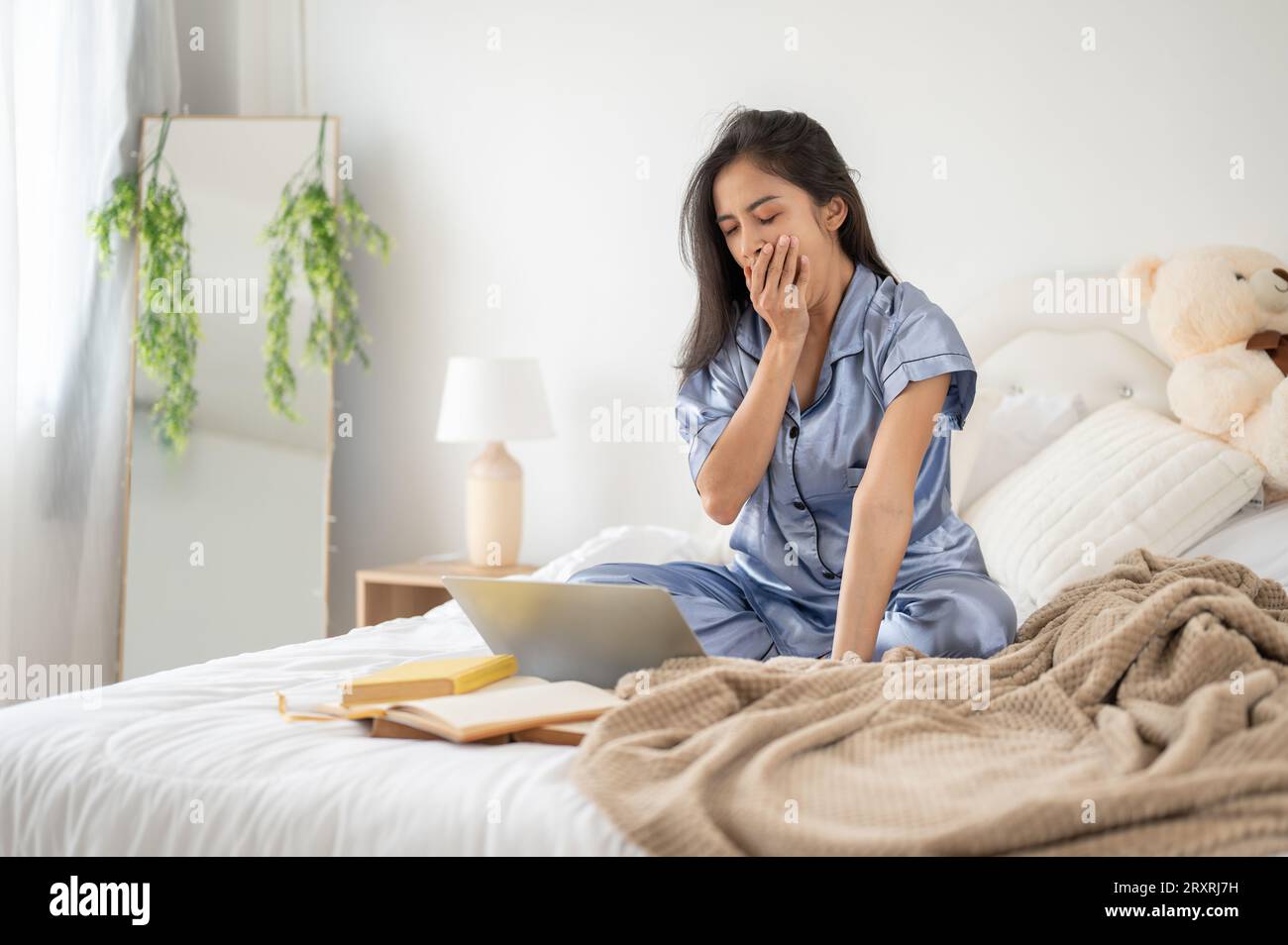 A sleepy and tired Asian woman in pajamas is covering her mouth and yawning while working on her ...
