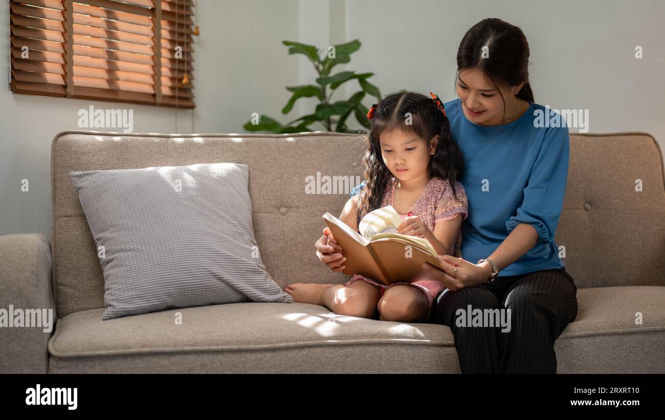 A happy and caring young Asian mom is reading a book, telling a story ...