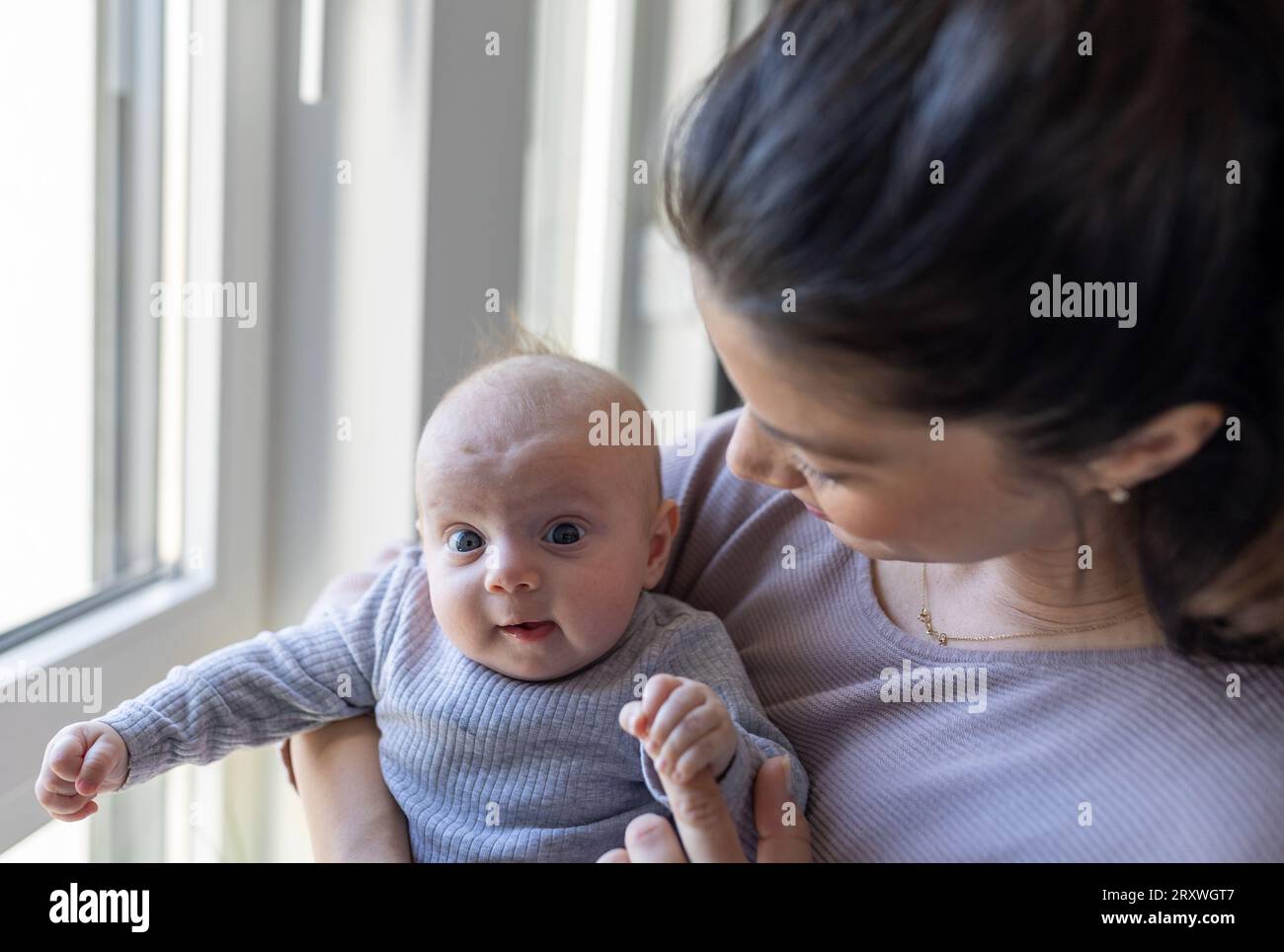 Happy young mother standing beside window and holding cute smiling ...