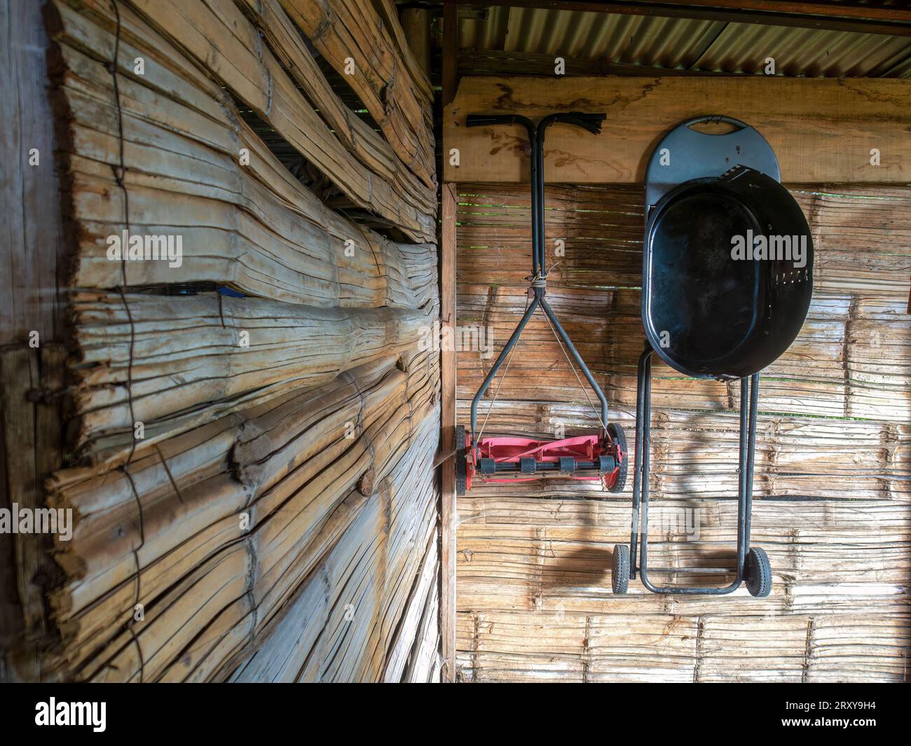 A manual lawn mower and a basic black grill hanging from a cane wall in ...