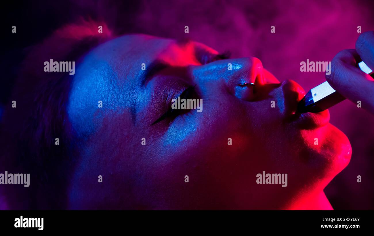 Portrait asian woman with shaved temples smokes vape in neon light ...