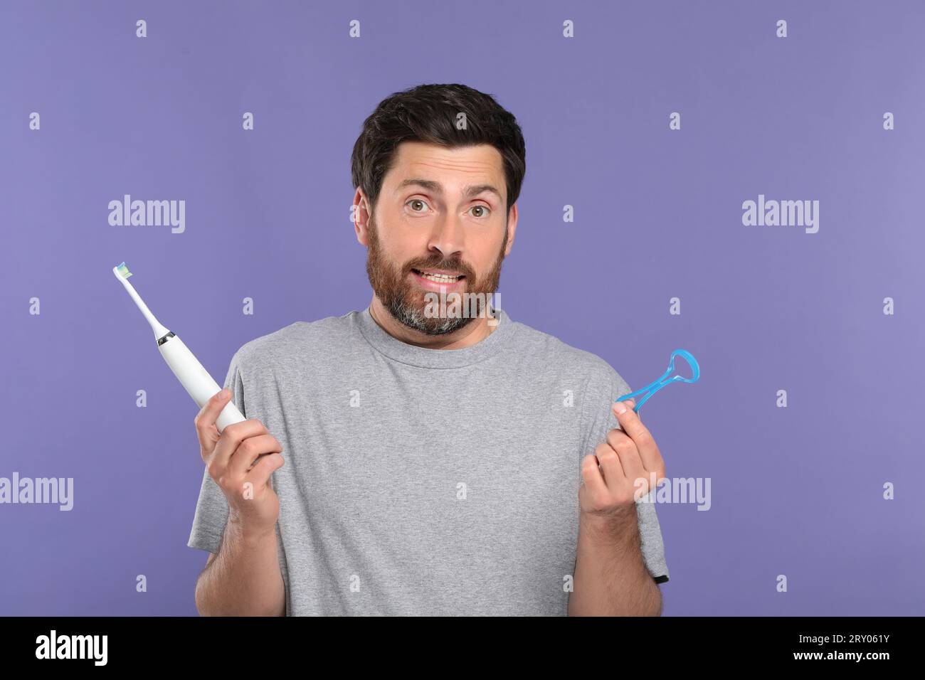 Confused man with tongue cleaner and electric toothbrush on violet ...