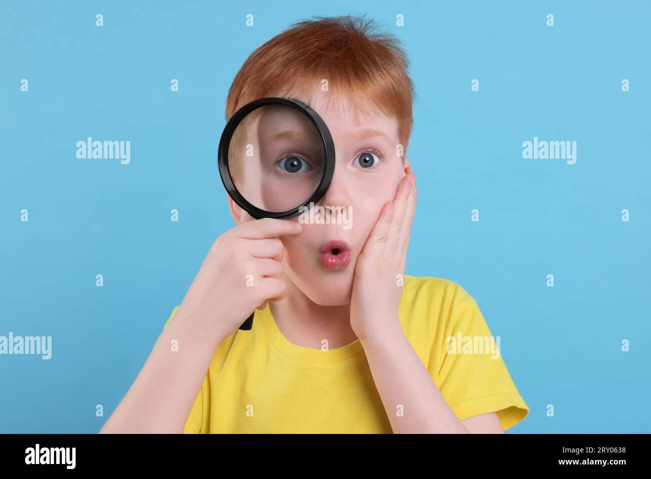 Surprised boy looking through magnifier glass on light blue background ...