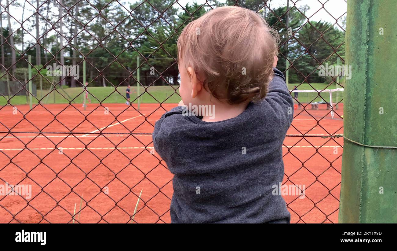 Cute baby holding into tennis court fence looking at siblings play game ...