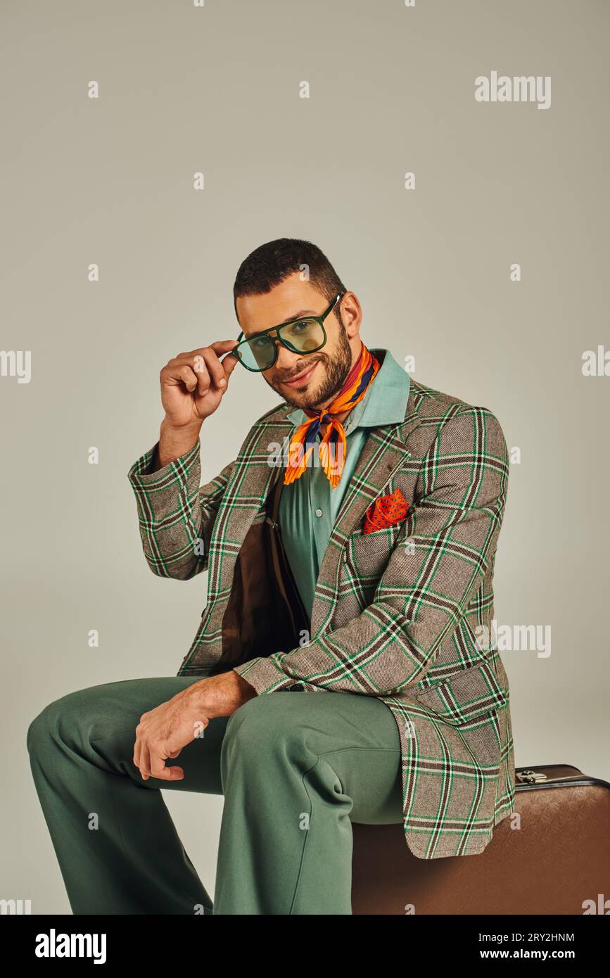 happy man in retro attire sitting on vintage suitcase and smiling at ...