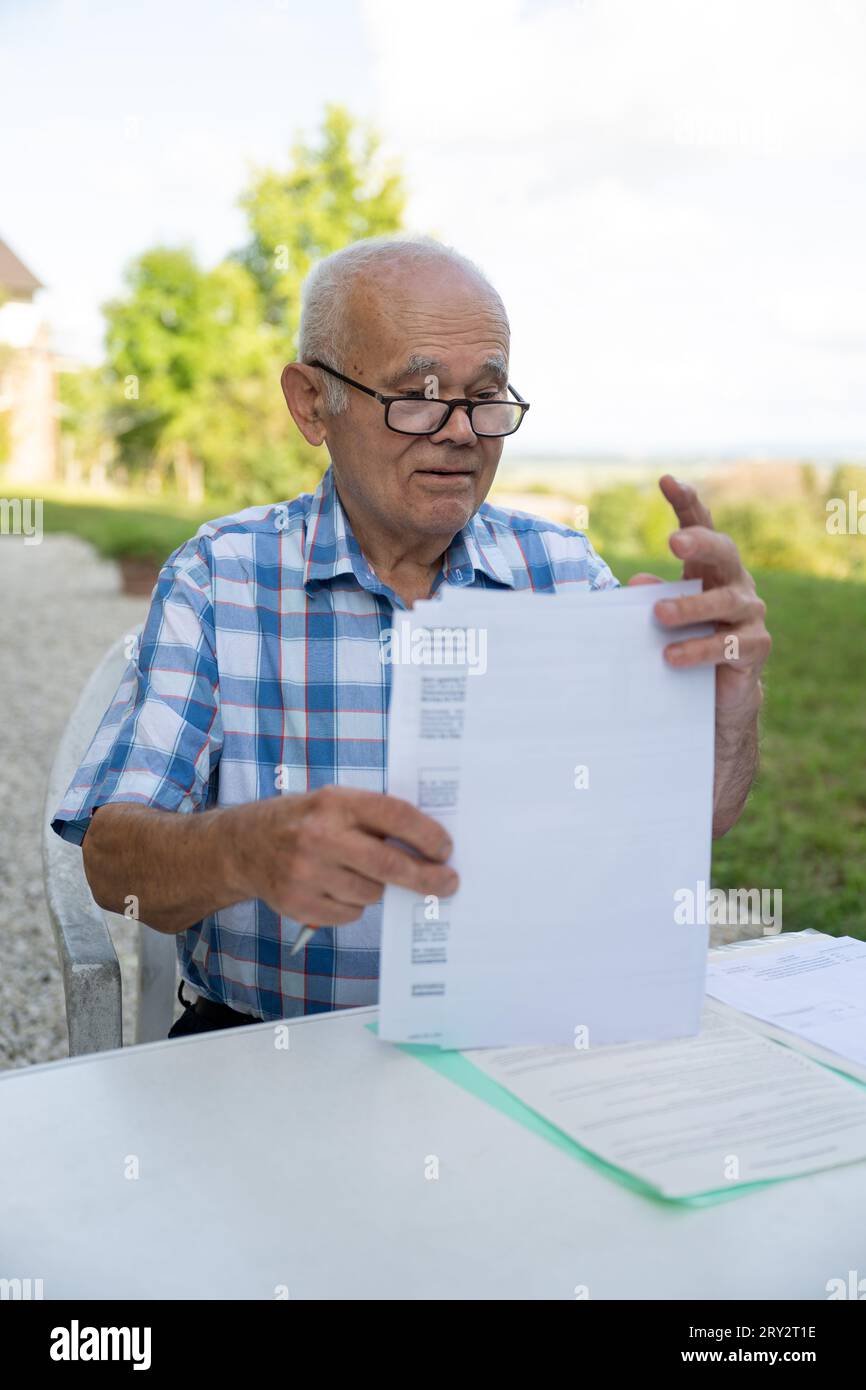 An elderly man sits at a desk outside, working with documents, filling ...