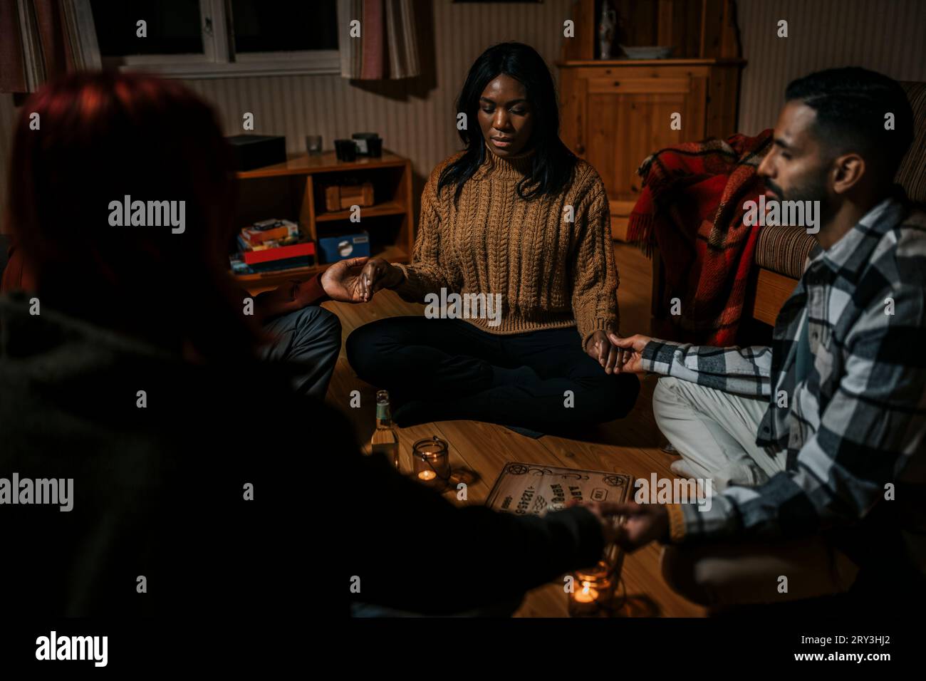 Multiracial friends holding hands sitting with ouija board in log cabin ...