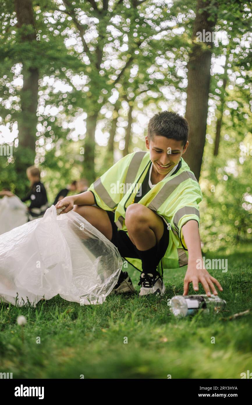 Full length of smiling boy crouching and collecting garbage in bag on ...