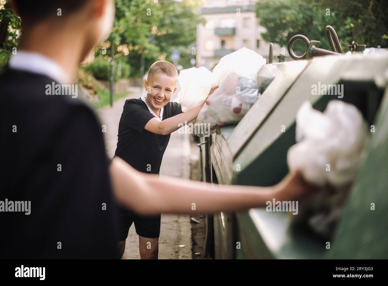 Smiling boy looking at friend while putting garbage bags in recycling ...